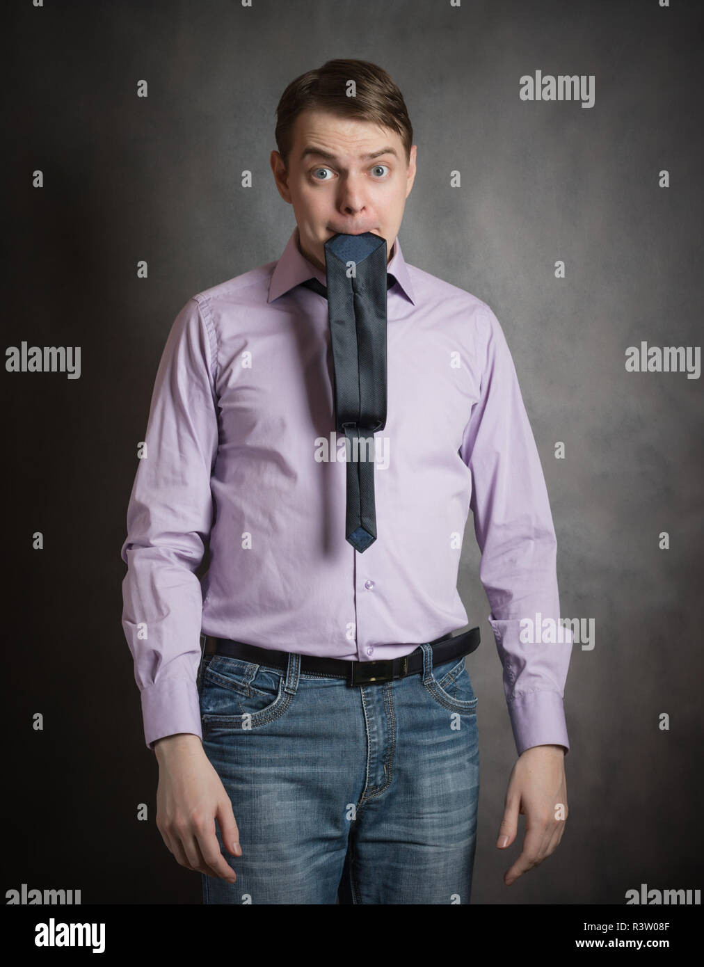 Emotional young man in pink shirt biting tie. Against dark background ...