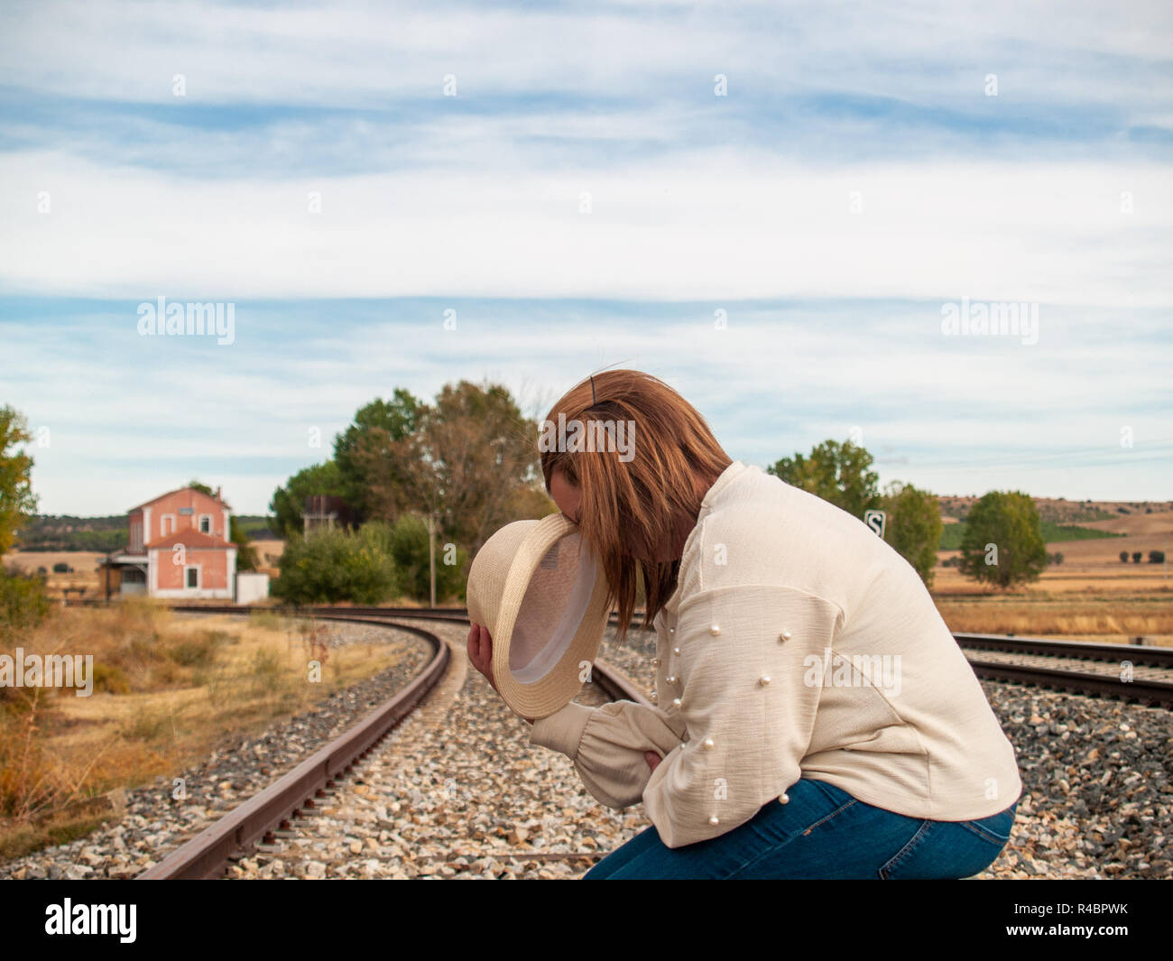 A sad blonde woman with a straw hat on the train tracks sitting on a ...