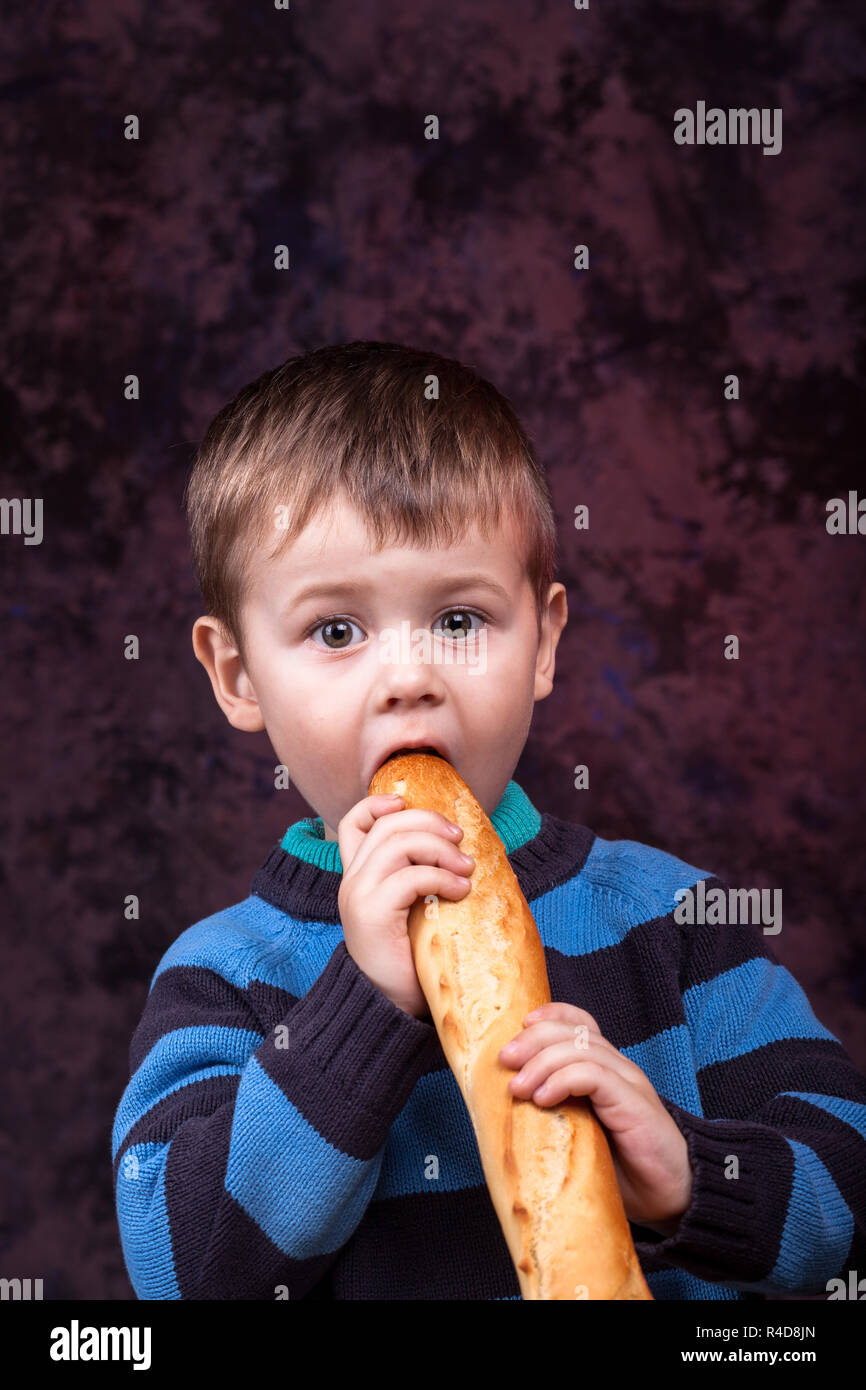 Cute kid holding and biting French bread against dark red background ...