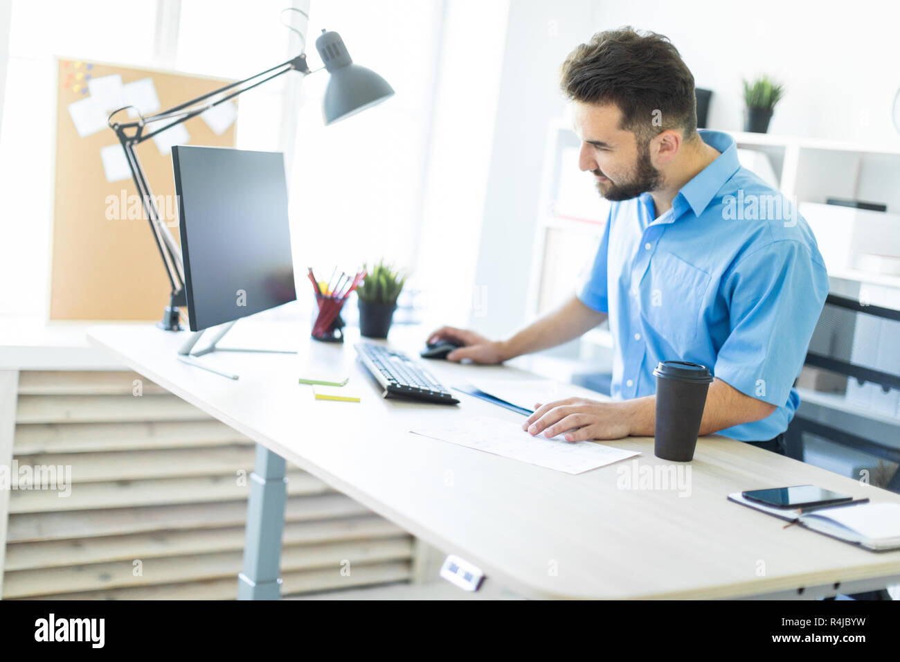 A young man standing in the office and working at the computer table ...