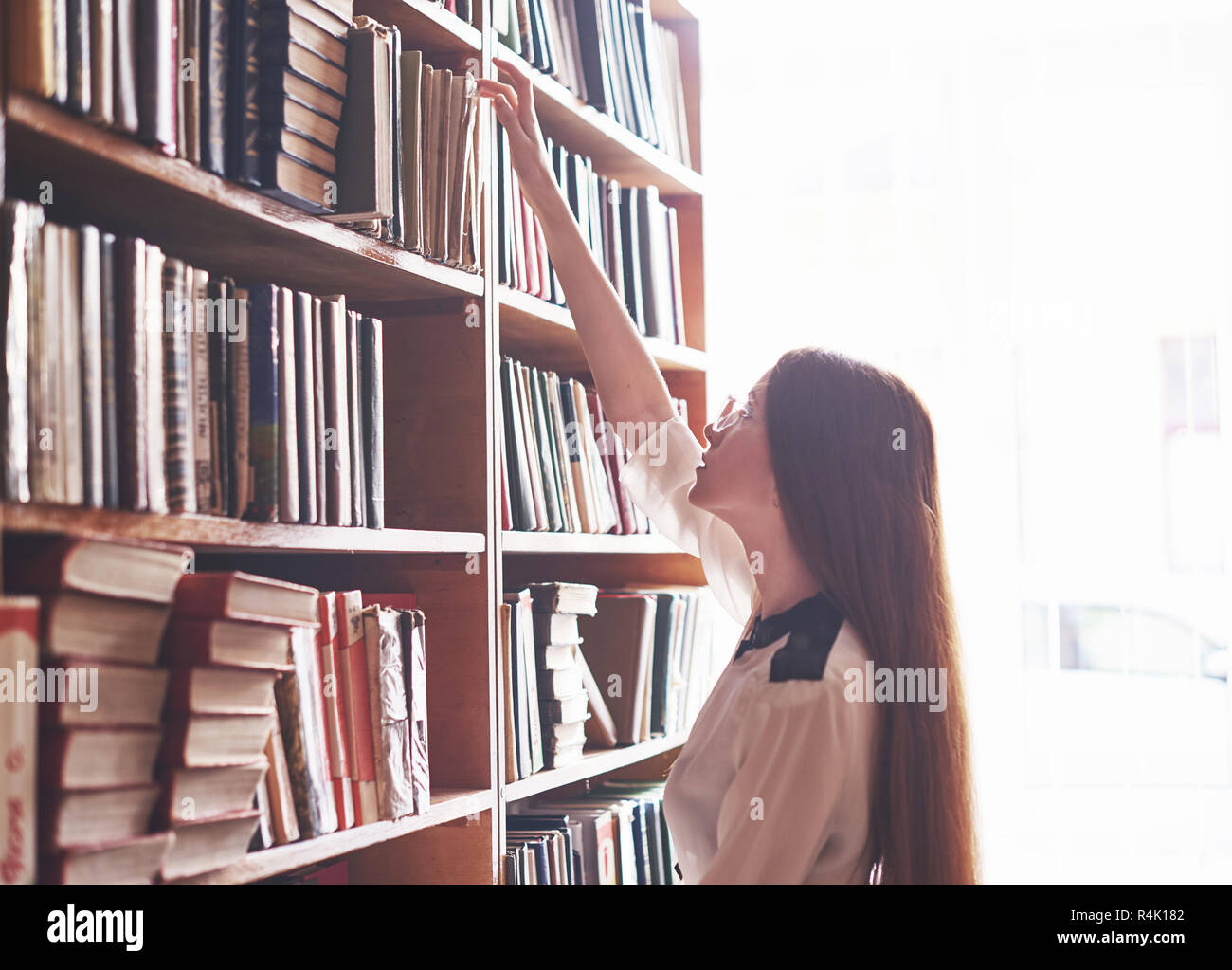 A young student girl is looking for the right book on the shelves of ...