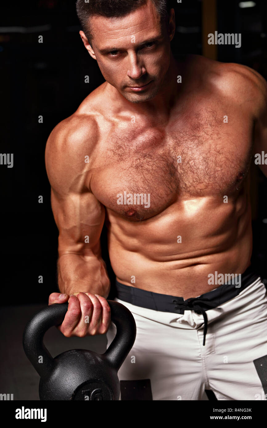 Strong angry athletic man doing the exercises with kettlebells on dark ...