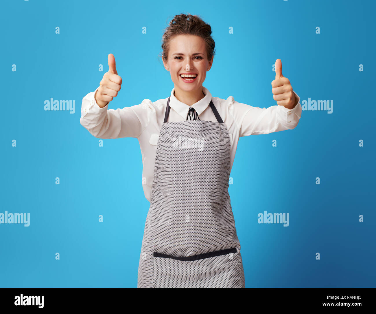smiling young cleaning lady in apron showing thumbs up against blue ...