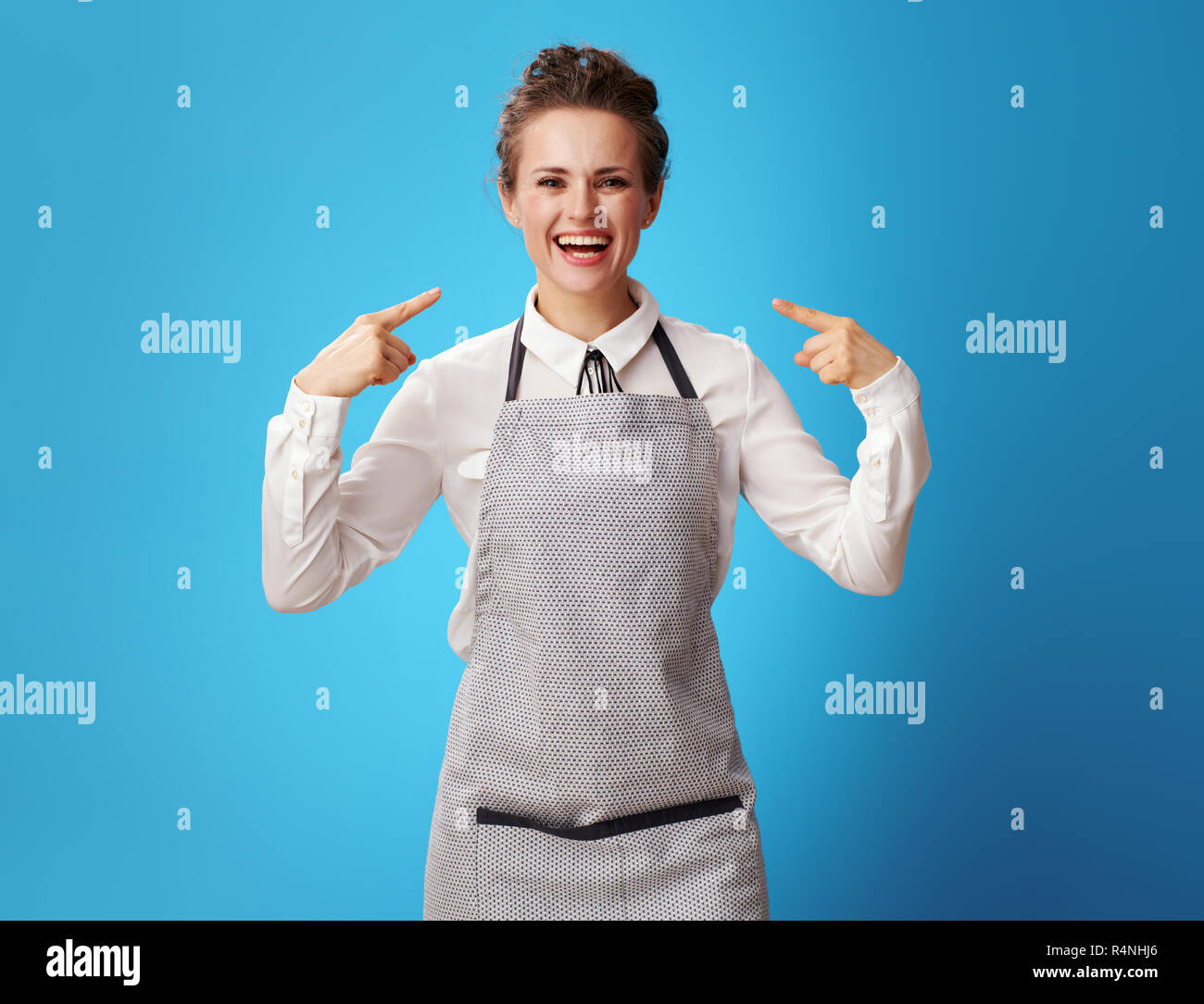 happy young cleaning lady in apron pointing at herself against blue ...
