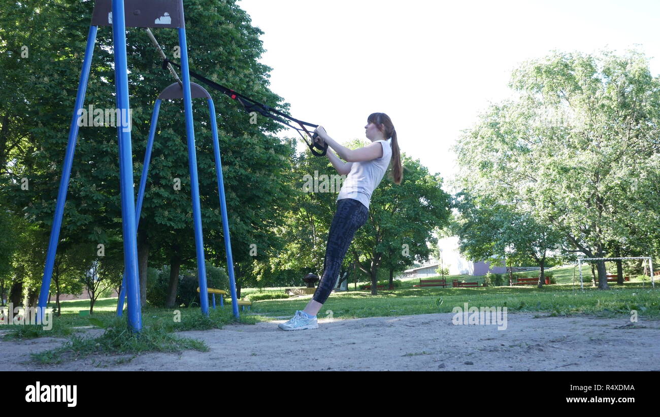 young woman doing intense pull-up exercising on fitness loops in summer ...