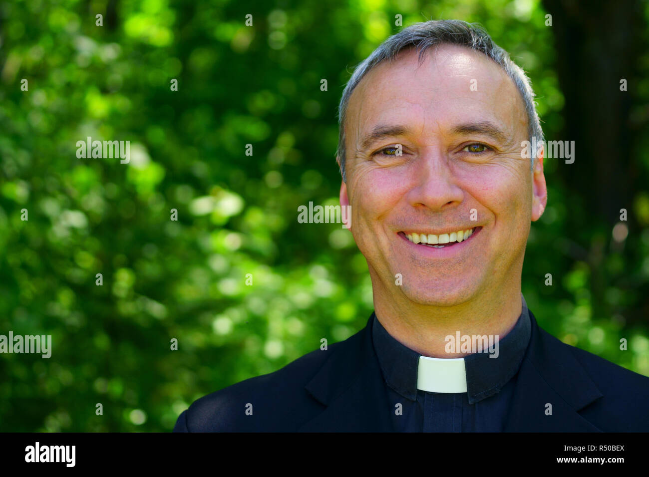 A good looking catholic priest is standing in the forest, smiling at us ...