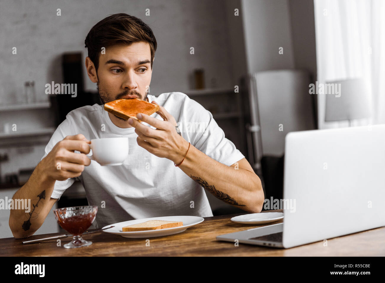 emotional young freelancer eating toast with jam and coffee and looking ...
