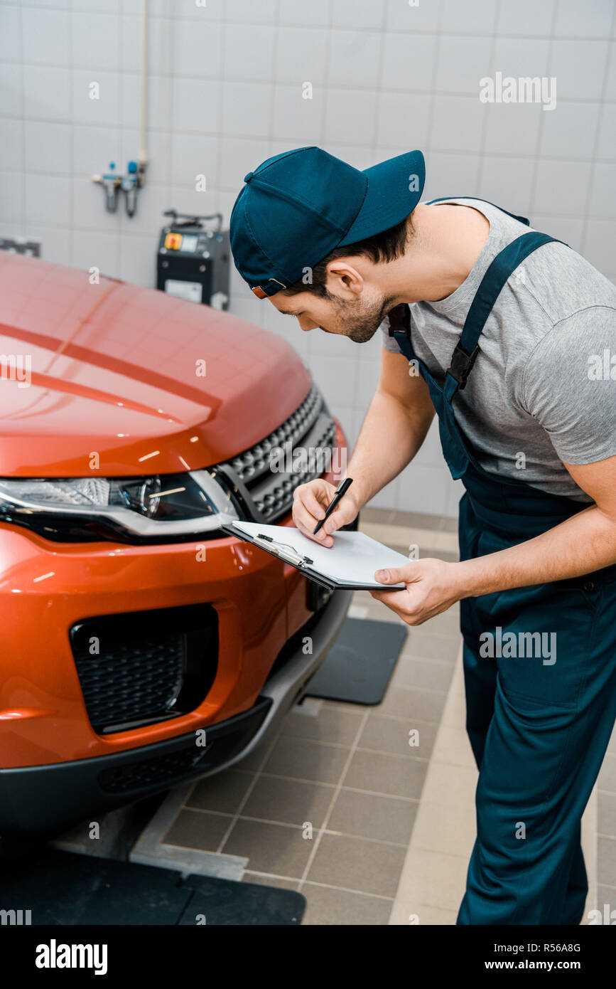young auto mechanic in uniform with notepad examining car at auto ...