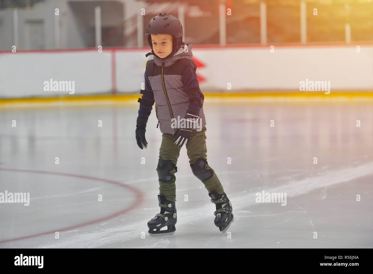 Adorable little boy in winter clothes with protections skating on ice ...