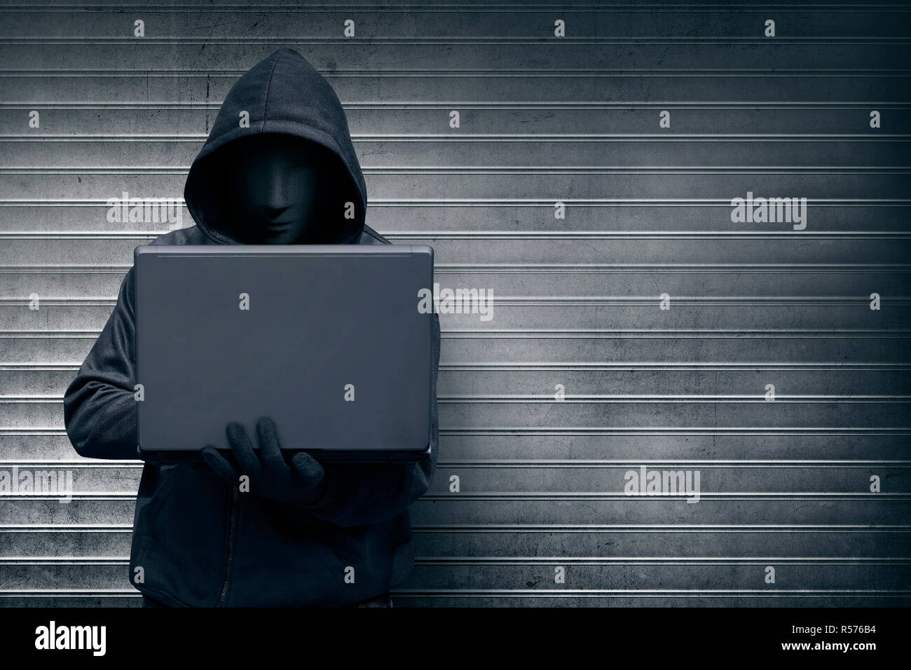 Hooded hacker with mask holding laptop while typing Stock Photo - Alamy