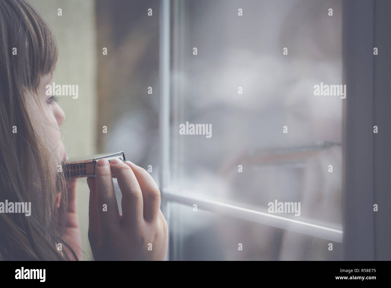 Sad Caucasian girl standing by the window and playing harmonica at home ...