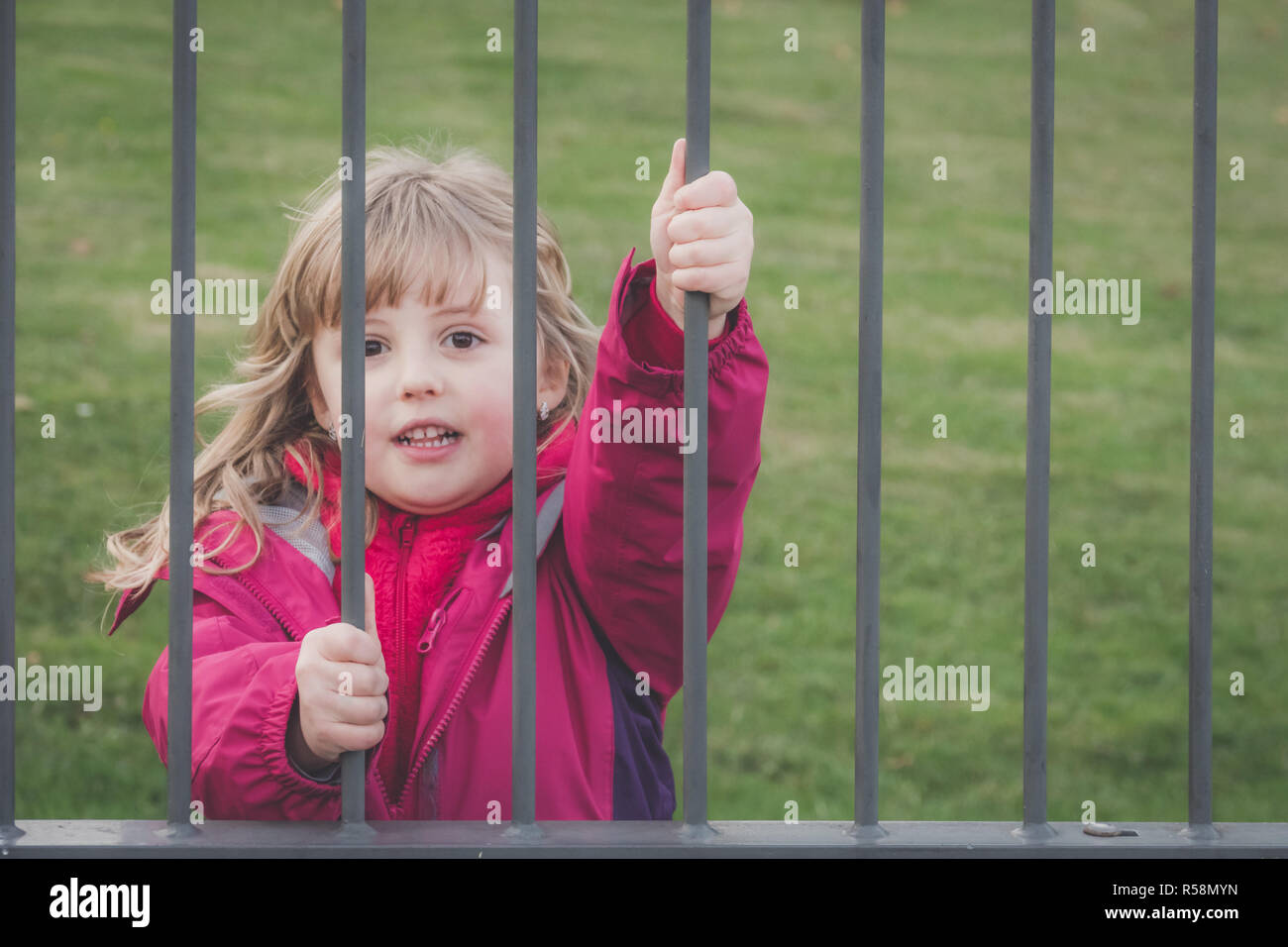 Little cute Caucasian girl dressed in pink jacket standing behind metal ...