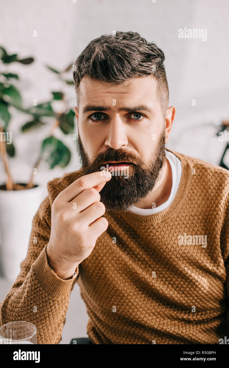 portrait of sick bearded man looking at camera and taking pill from ...