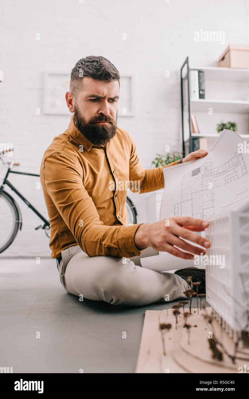 focused bearded adult male architect holding blueprint and working on ...