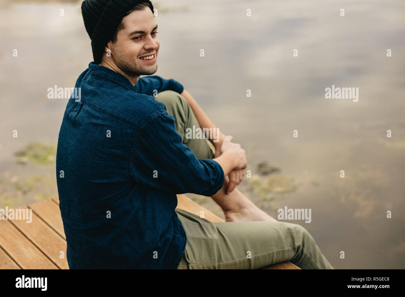 Smiling tourist man sitting on a wooden dock on a lake in a relaxed ...