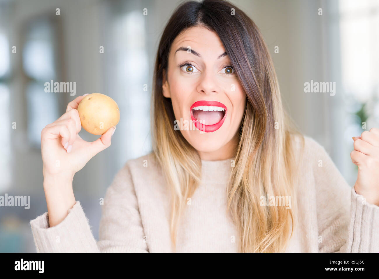 Young beautiful woman holding fresh potato at home screaming proud and ...