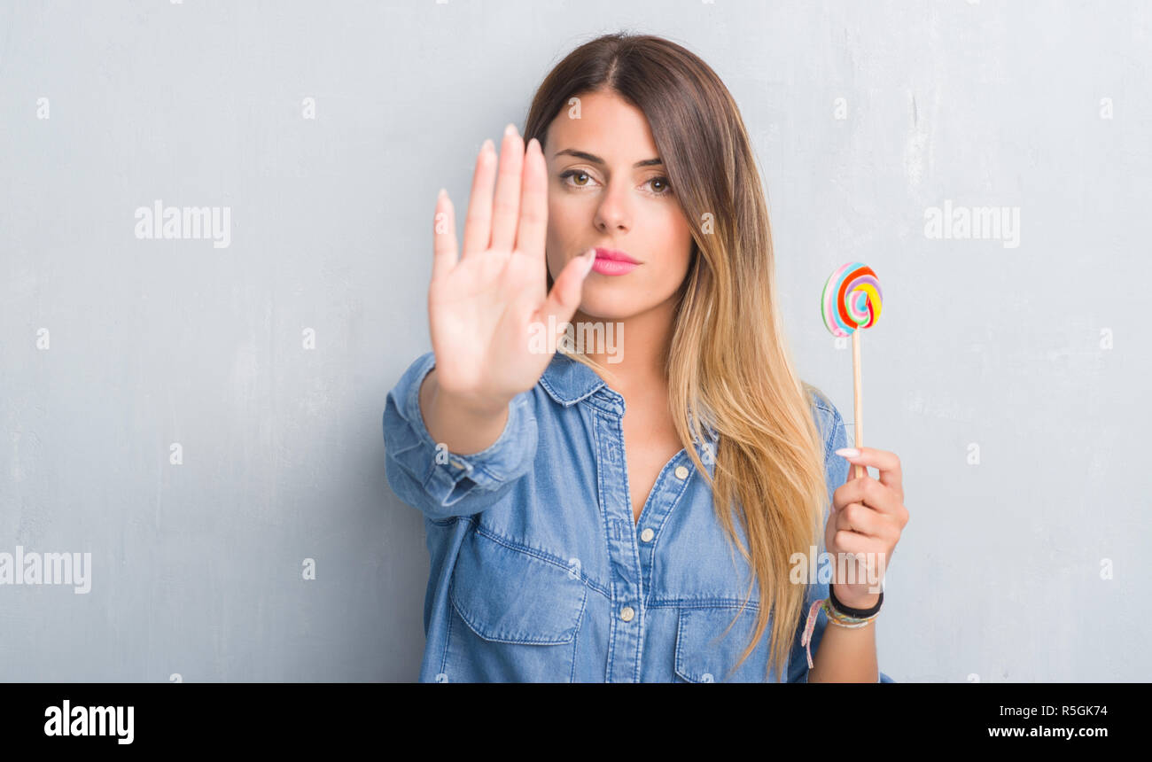 Young adult woman over grey grunge wall eating lollipop candy with open ...