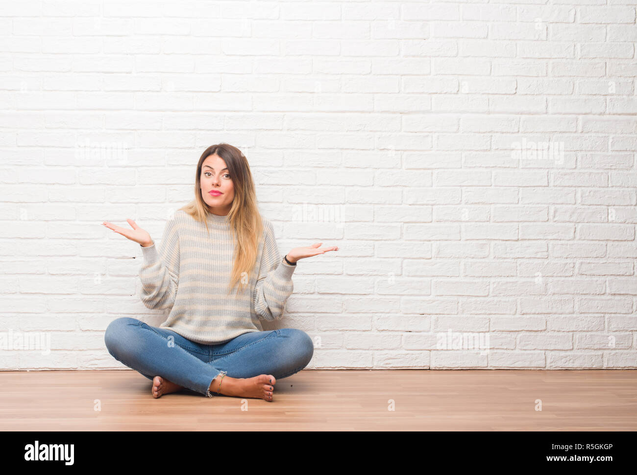 Young adult woman sitting on the floor over white brick wall at home ...