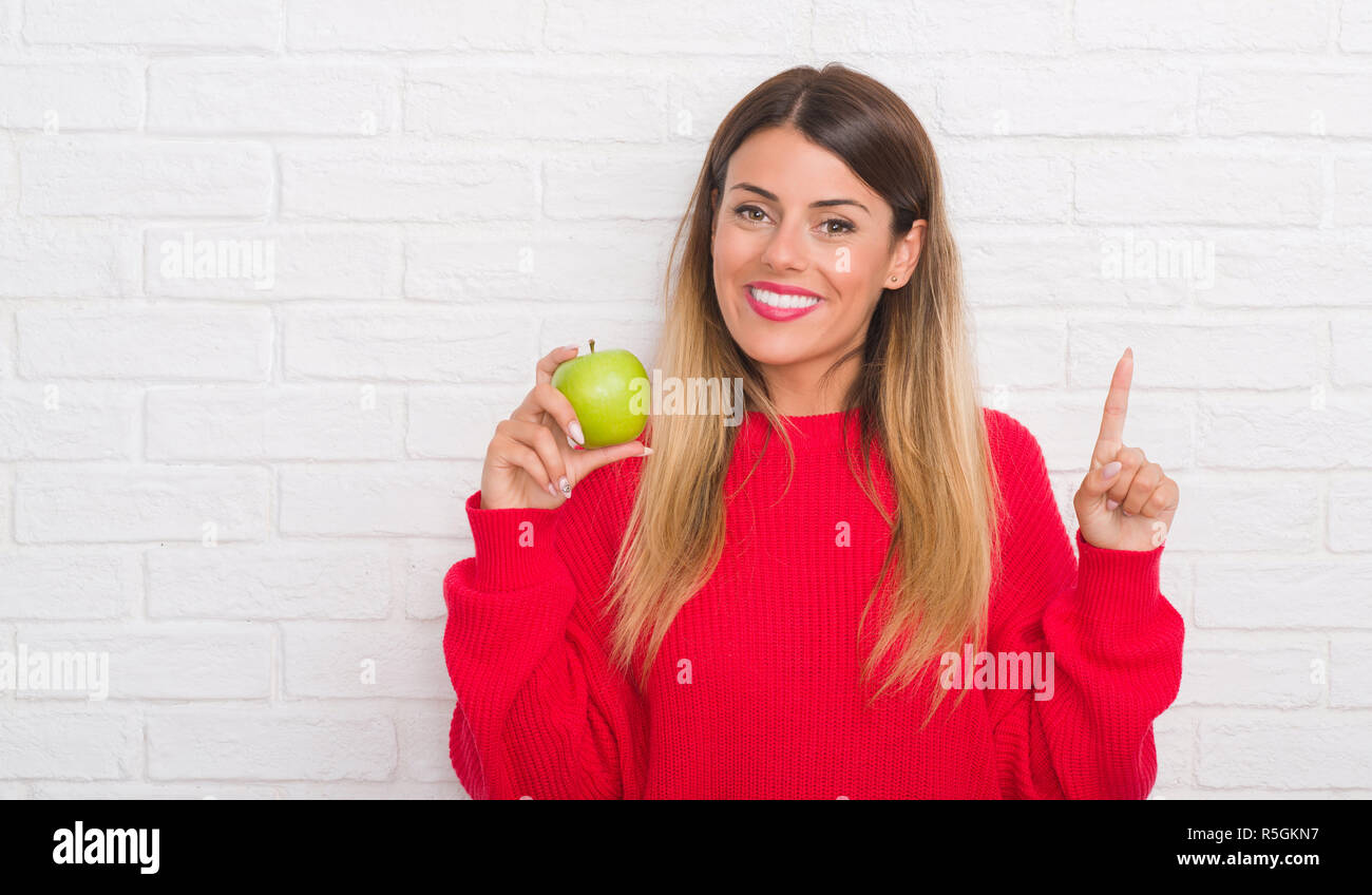 Young adult woman over white brick wall eating fresh green apple ...
