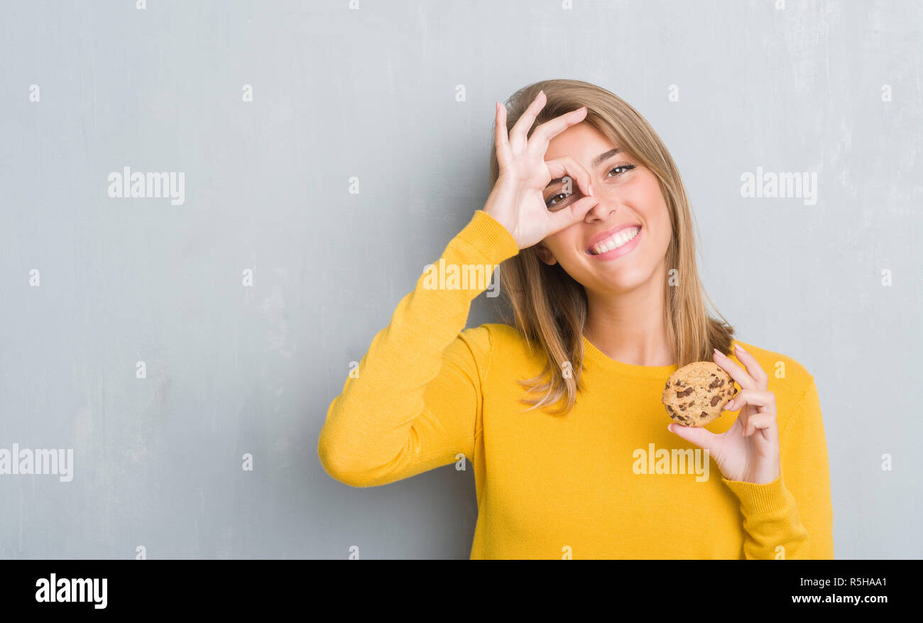 Beautiful young woman over grunge grey wall eating chocolate chip cooky ...