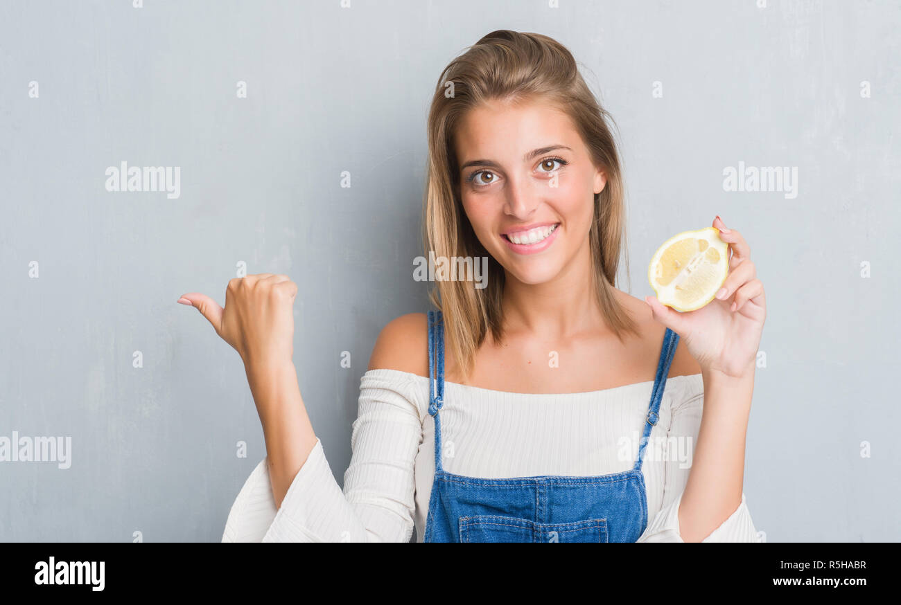 Beautiful young woman over grunge grey wall holding a lemon pointing ...