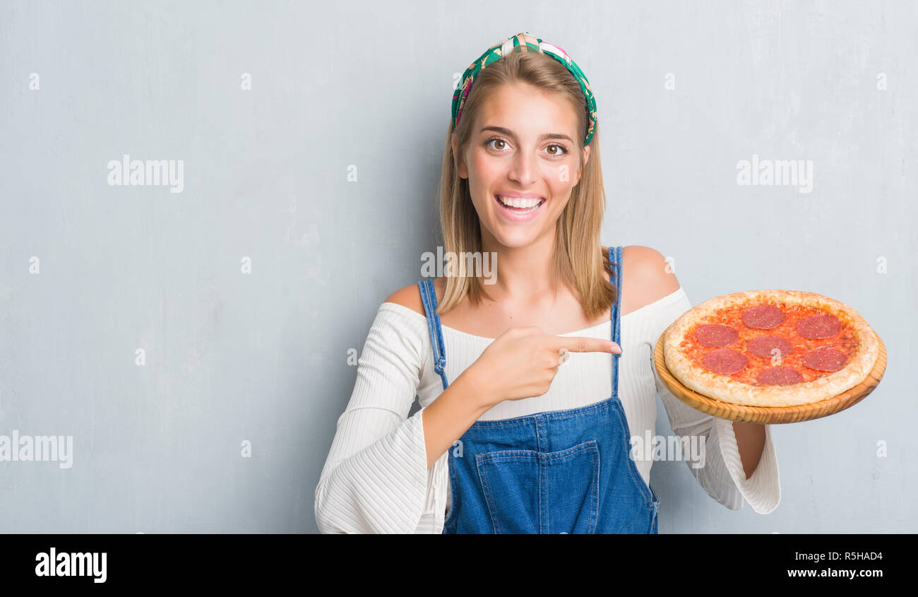 Beautiful young woman over grunge grey wall eating pepperoni pizza very ...