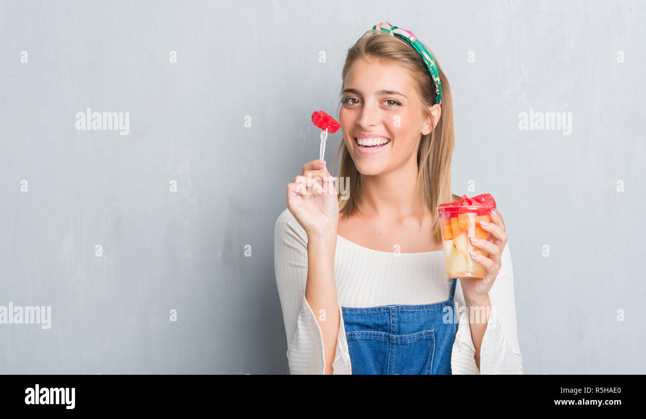 Beautiful young woman over grunge grey wall eating fruits with a happy ...