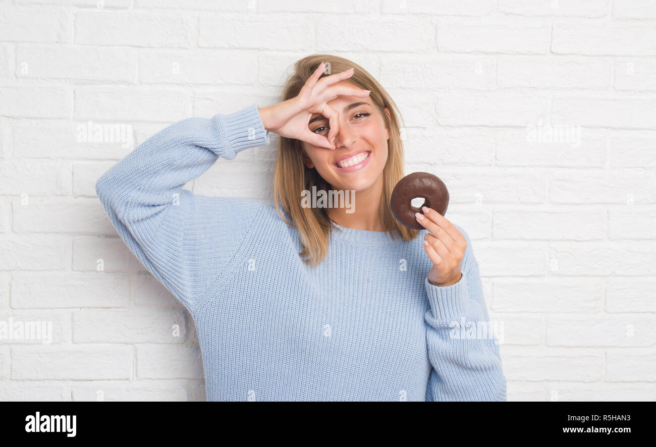 Beautiful young woman over white brick wall eating chocolate donut with ...