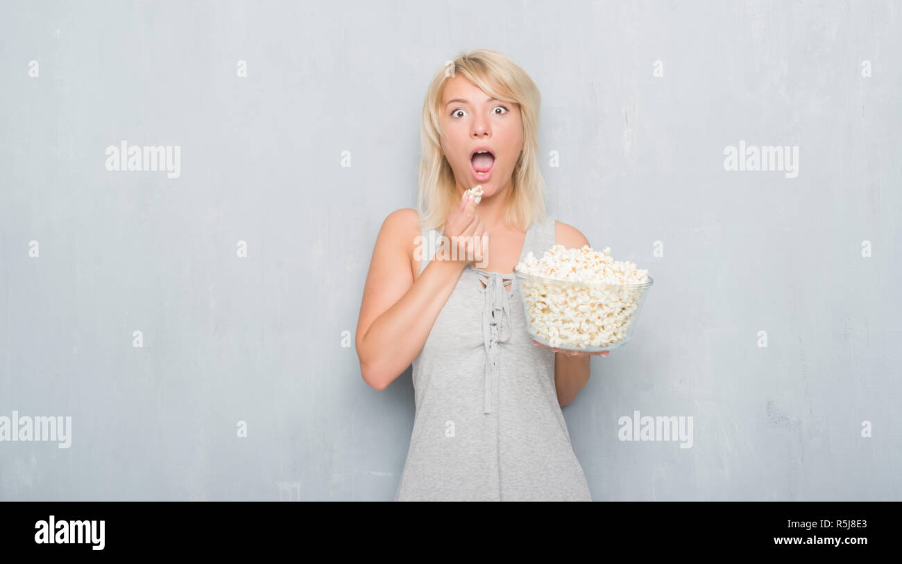Adult caucasian woman over grunge grey wall eating pop corn scared in ...