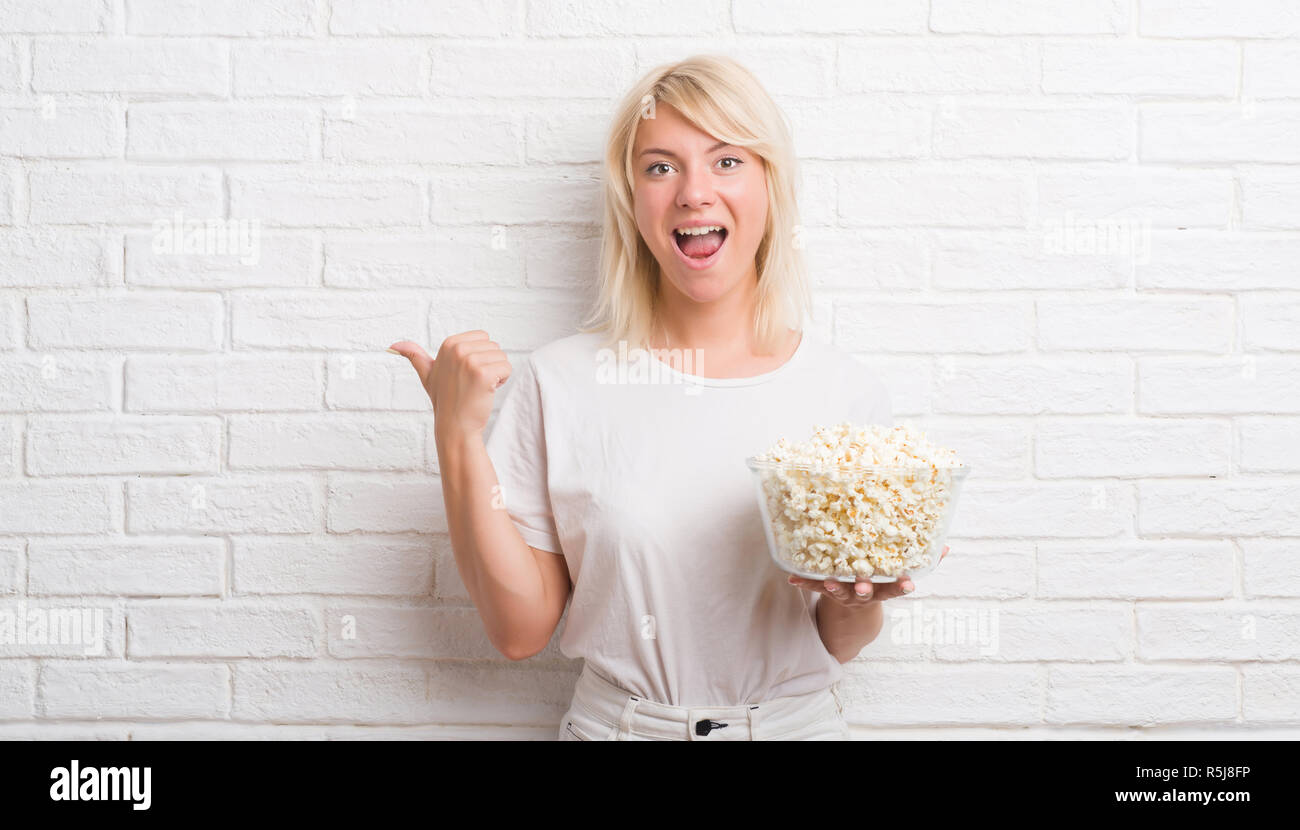 Adult caucasian woman over white brick wall eating pop corn pointing ...
