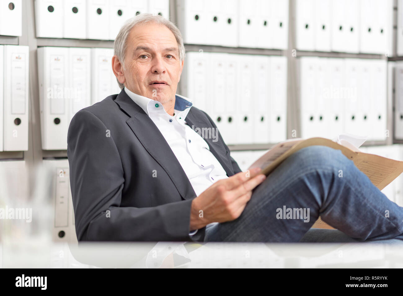 senior man in suit sitting in front of a shelf wall with documents ...