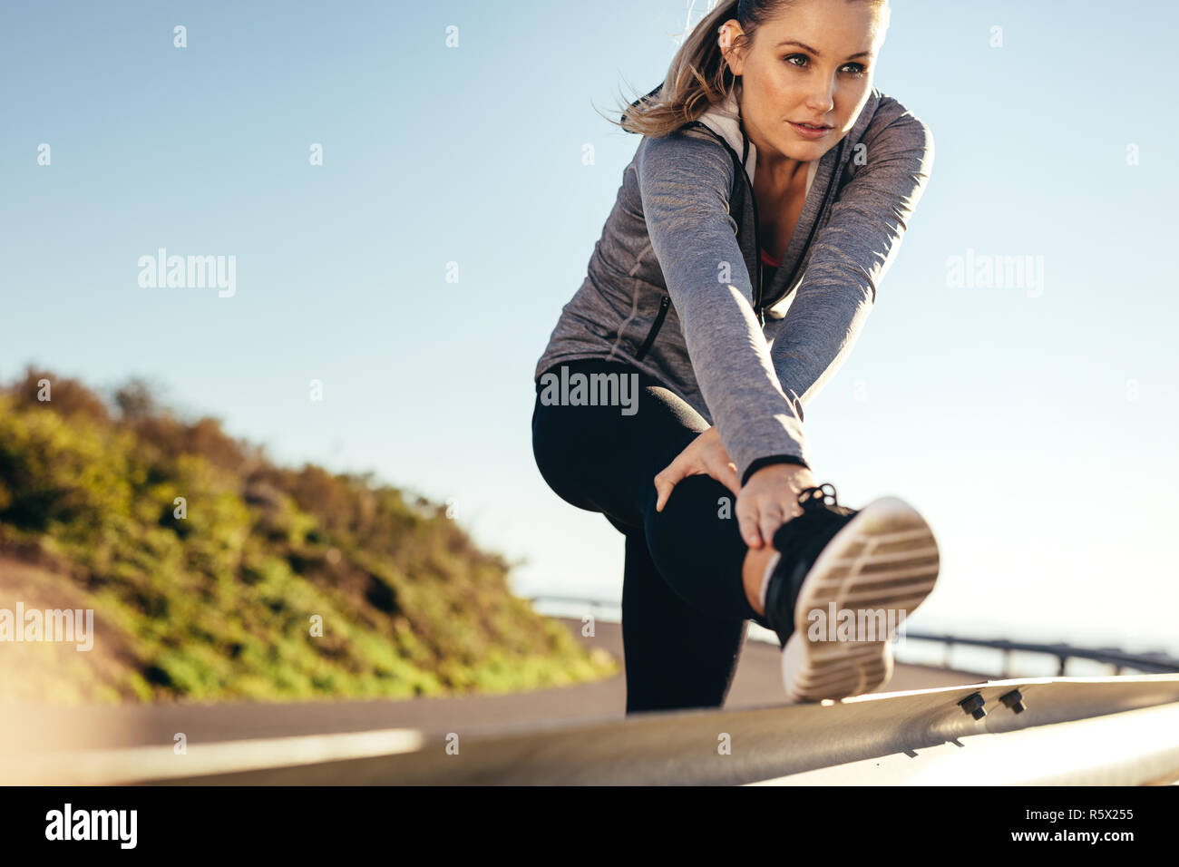 Woman athlete doing stretching exercises on the side railing of a road ...