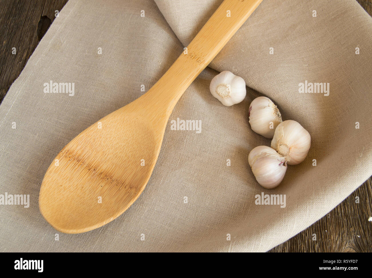 Garlic and a wooden spoon lying on a linen napkin. Old dark farm table ...
