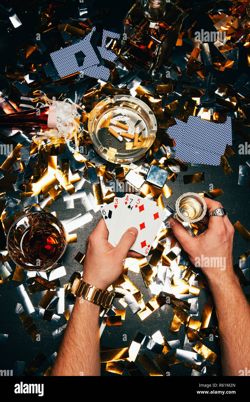 partial view of man with playing cards and alcohol sitting at table ...