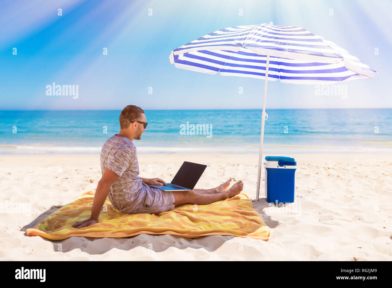 Young man lusing laptop computer on beach bench with blue umbrella ...