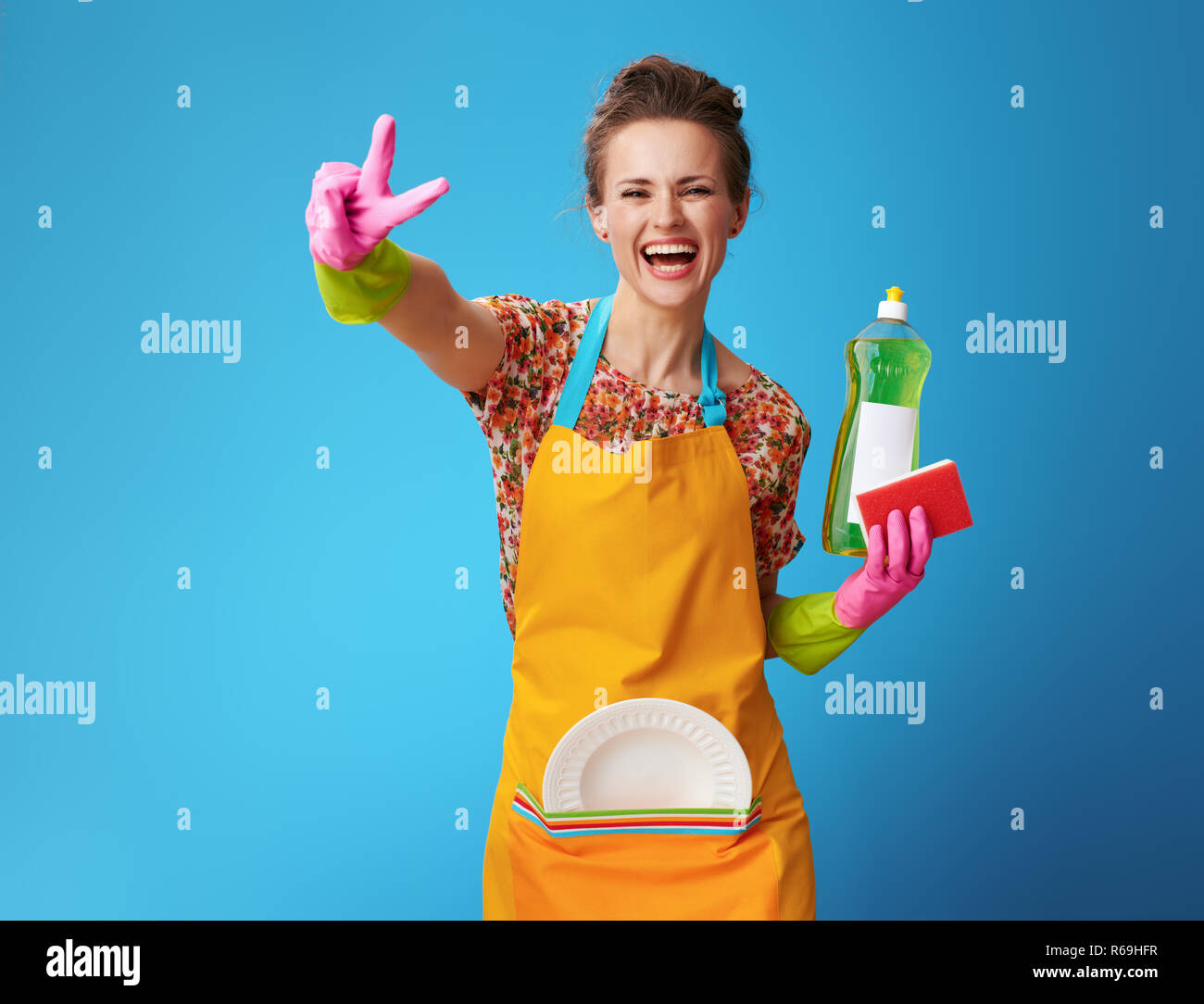 smiling young housewife in orange apron with sponge and dishwashing ...