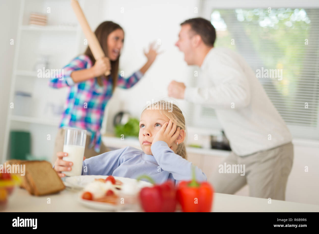 Beautiful little girl having breakfast in the kitchen and listening quarrel of parents. She is ...
