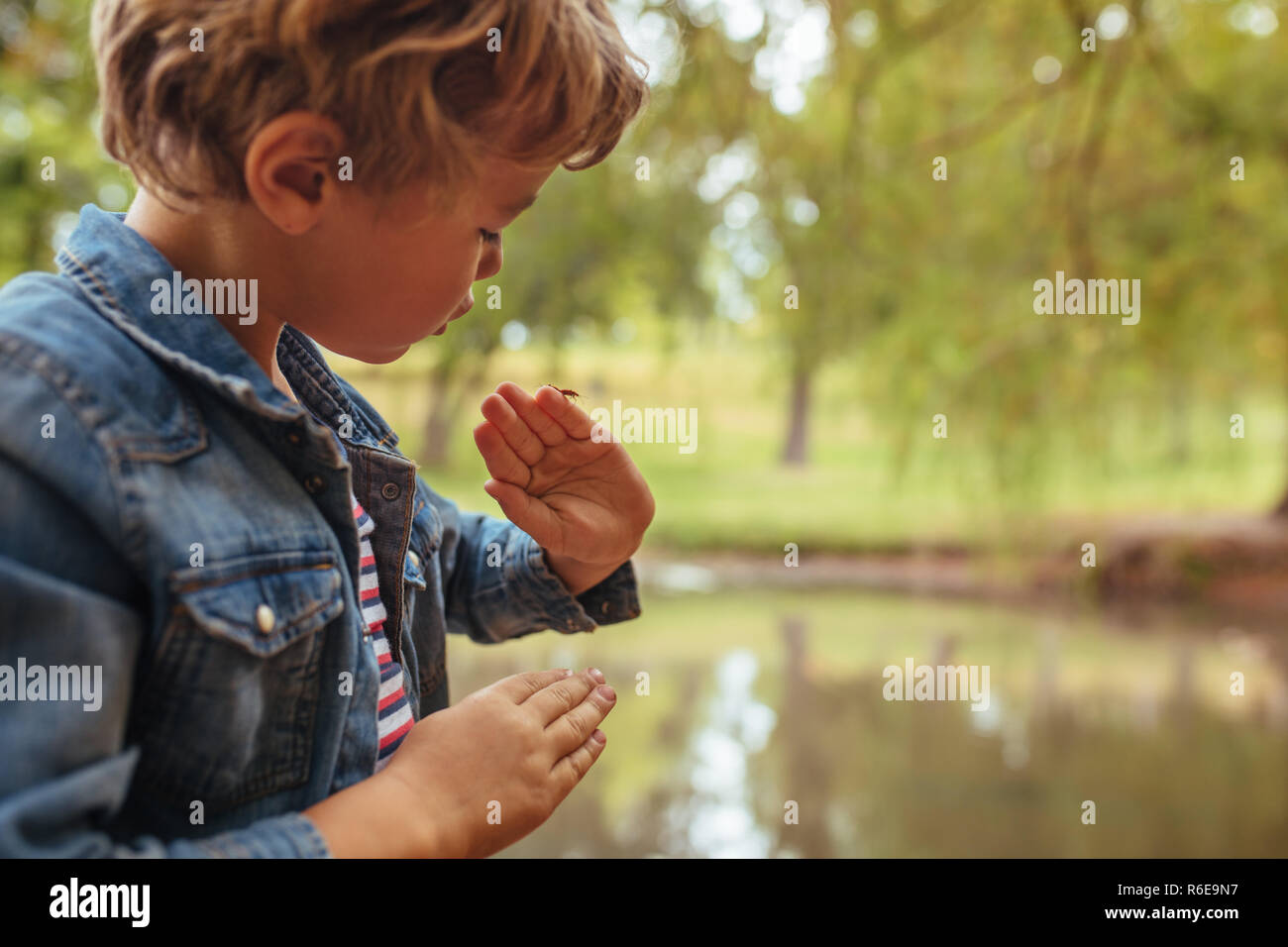 Little boy looking at an insect on his hand while standing by a pond in ...