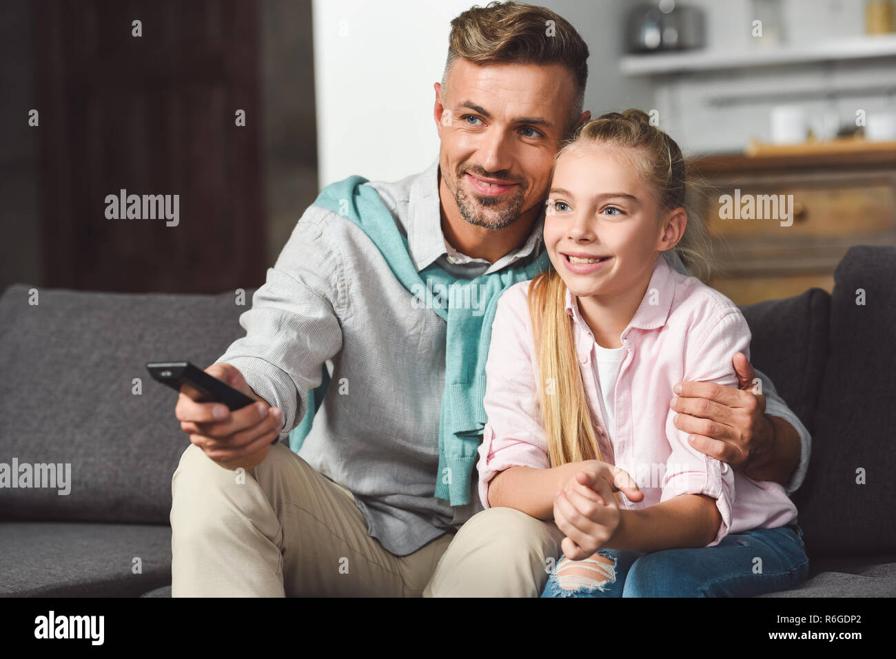 father with remote controller sitting on sofa and hugging daughter ...