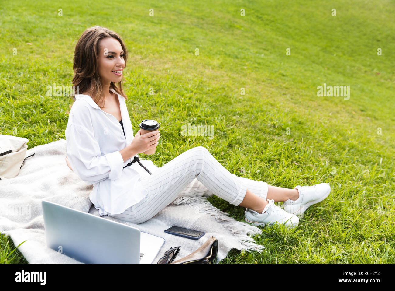 Smiling young woman spending time at the park, sitting on a grass with ...