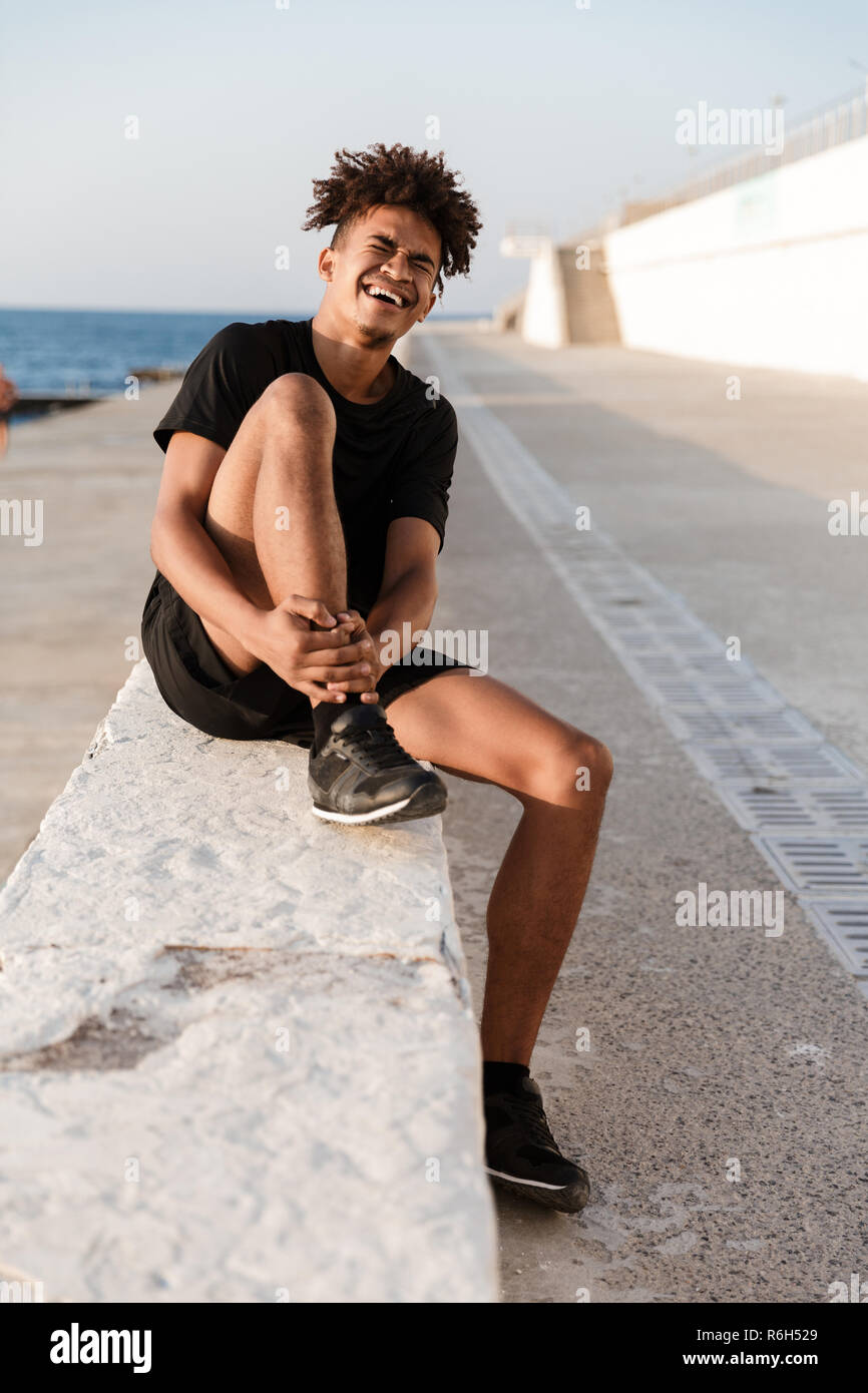 Image of sad young guy sportsman sitting with pain in leg outdoors on ...