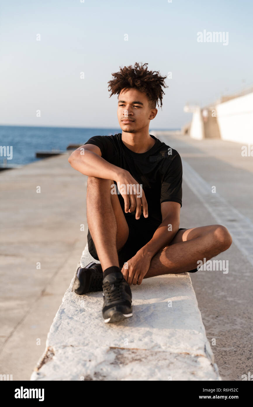 Image of concentrated young guy sportsman sitting outdoors on the beach ...