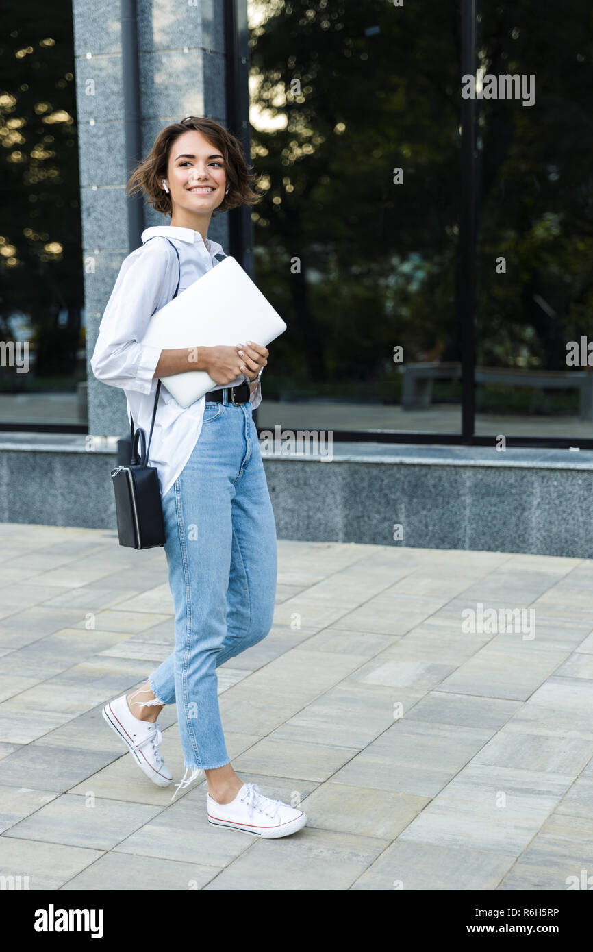 Smiling young woman walking outdoors at the street, carrying laptop ...