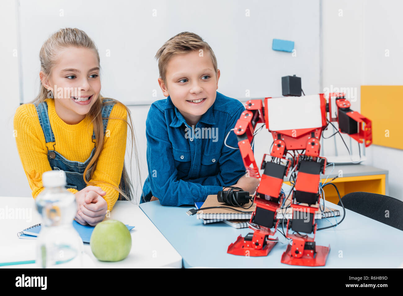 smiling schoolchildren sitting at desk and looking at red plastic robot ...