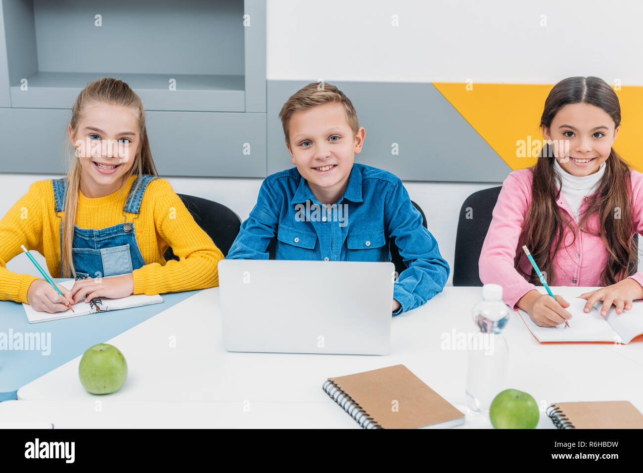 cute schoolchildren writing in notebooks, using laptop and looking at ...