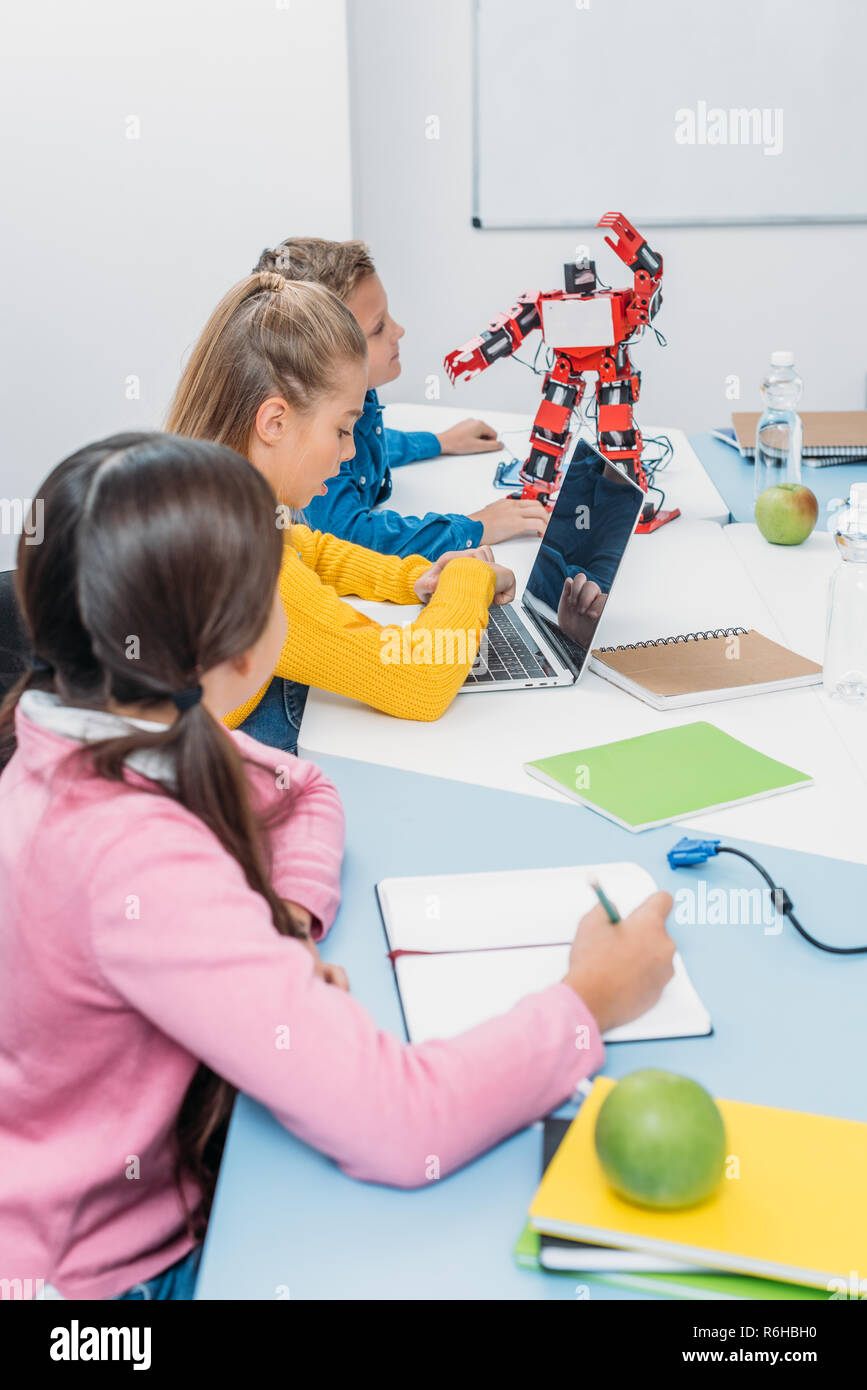 children writing in notebooks, using laptop and looking at red robot at ...