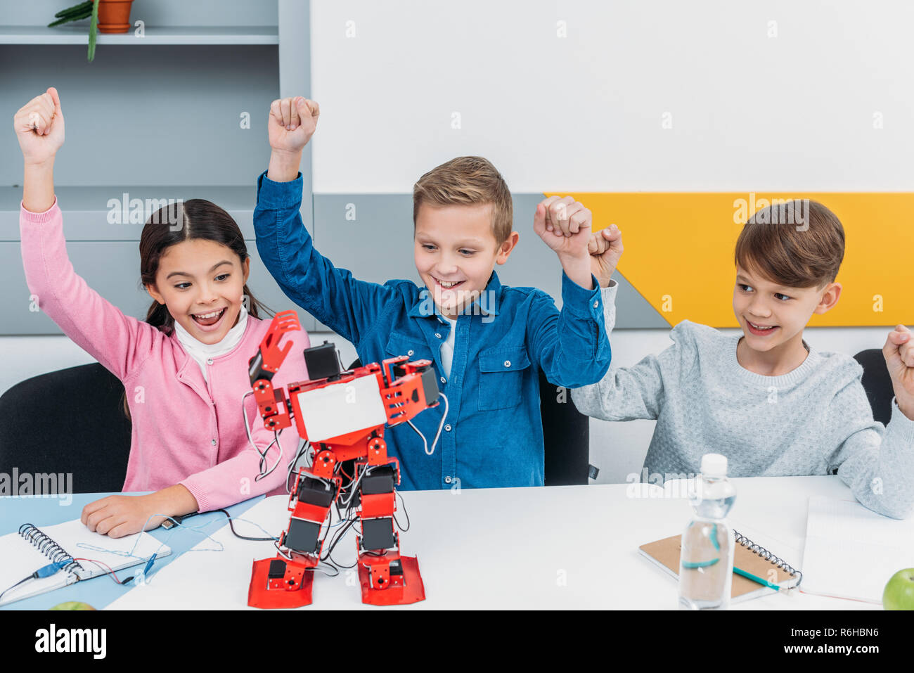 happy schoolchildren raising hands and looking at red handmade robot at ...