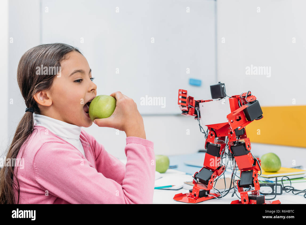 schoolgirl eating apple and sitting at table with robot model at STEM ...