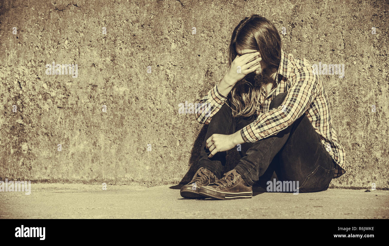 Man bearded long hair sitting sad alone by grunge wall outdoor ...