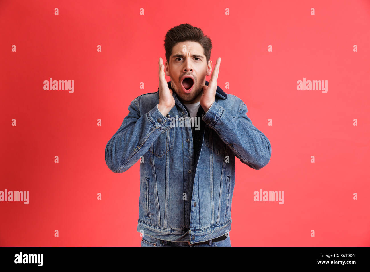 Portrait of a shocked young man dressed in denim jacket standing ...